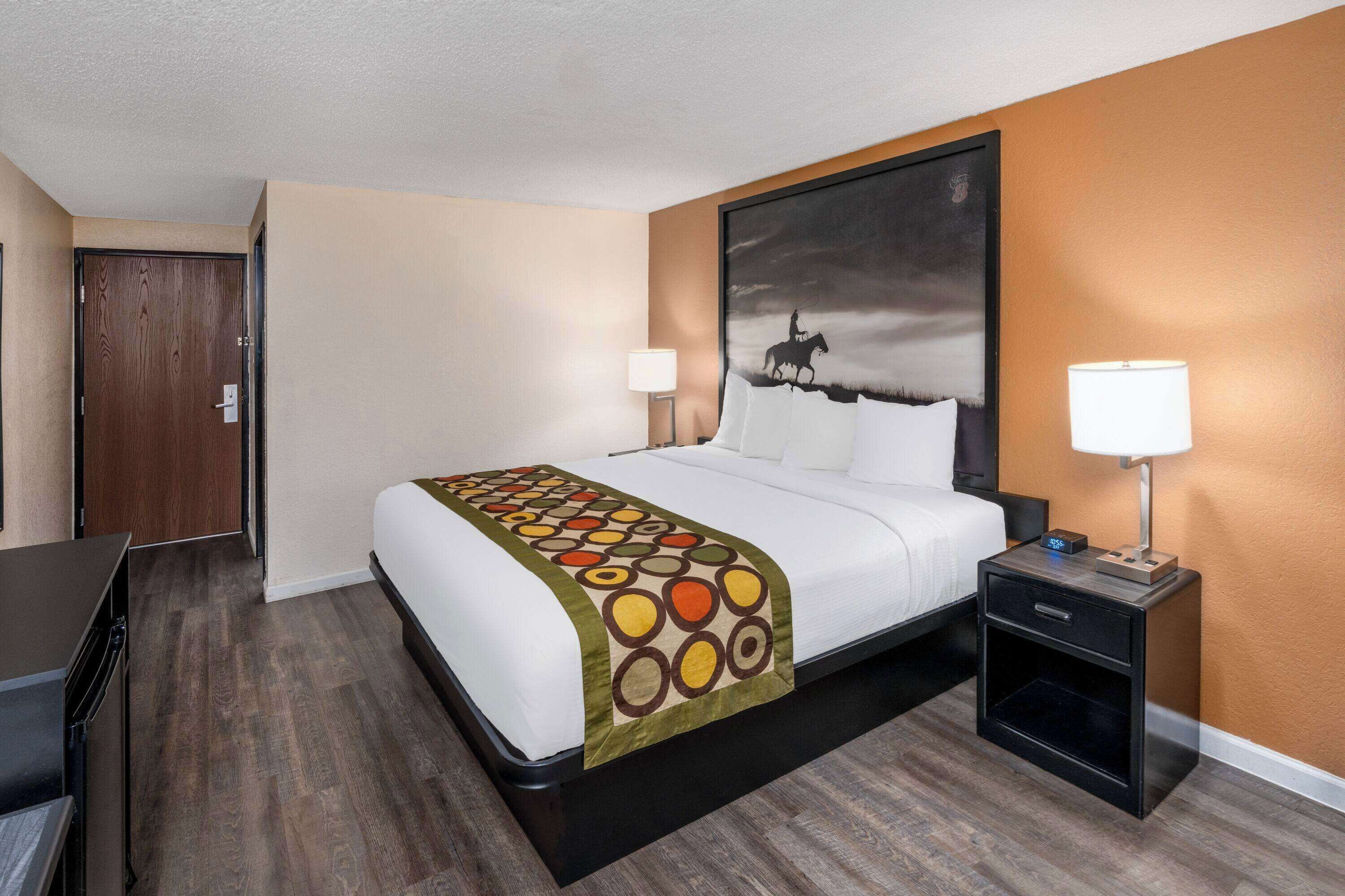 Quarto de hotel com cama, mesas de cabeceira, escrivaninha e um quadro na parede.