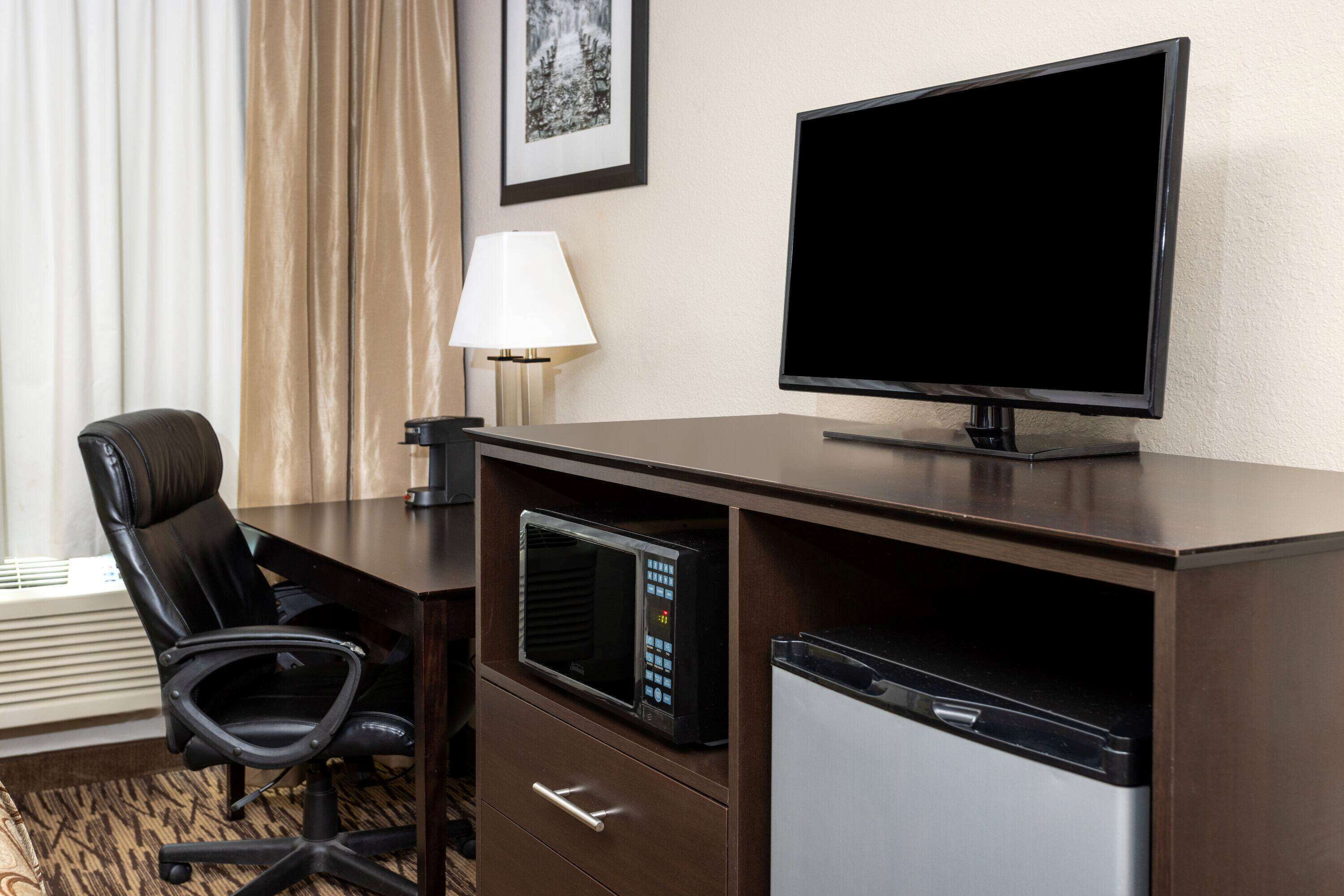 Quarto de hotel com escrivaninha, televisão, micro-ondas, frigorífico, cadeira, candeeiro e janela com cortinas.