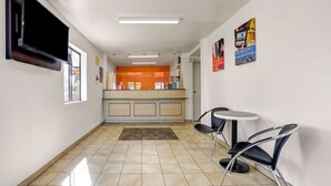 Um saguão de hotel com uma mesa de recepção, uma TV presa na parede e uma pequena área de estar com duas cadeiras e uma mesa.