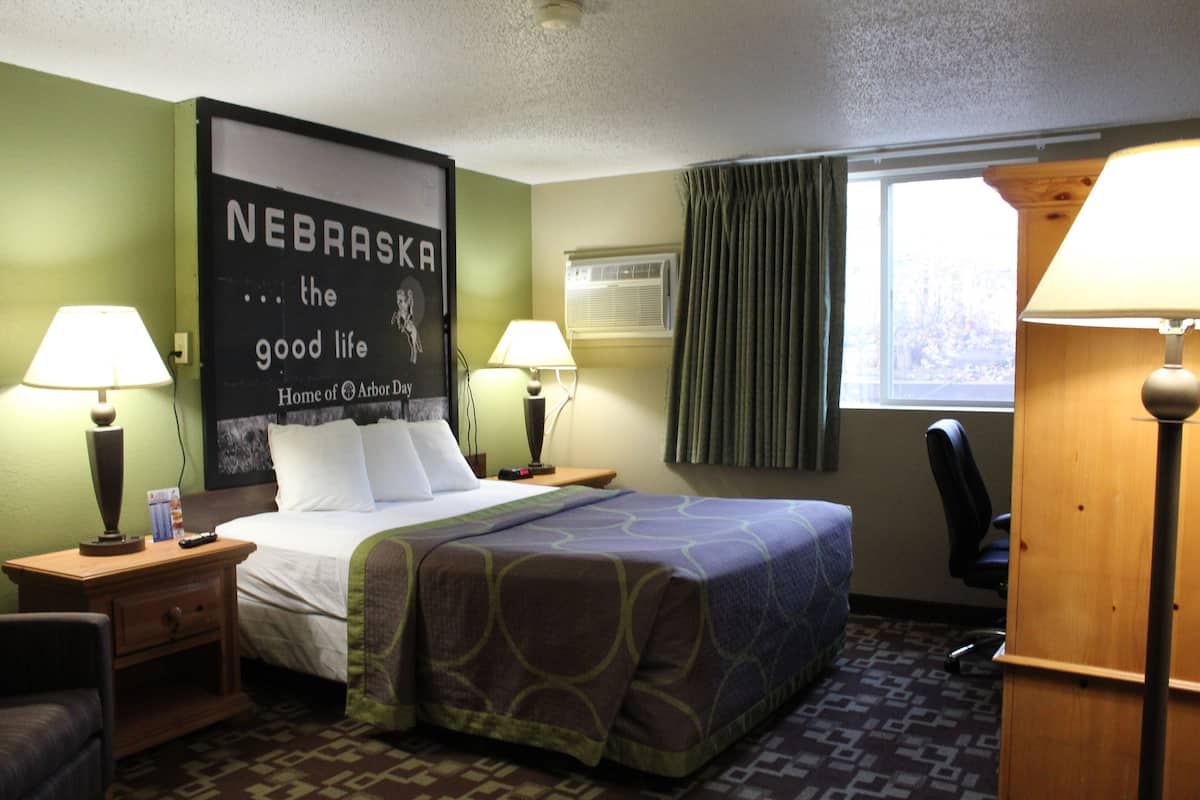 Quarto de hotel com uma cama, duas mesas de cabeceira com luminárias, uma escrivaninha, uma cadeira, uma janela com cortinas e uma parede com um letreiro de Nebraska.
