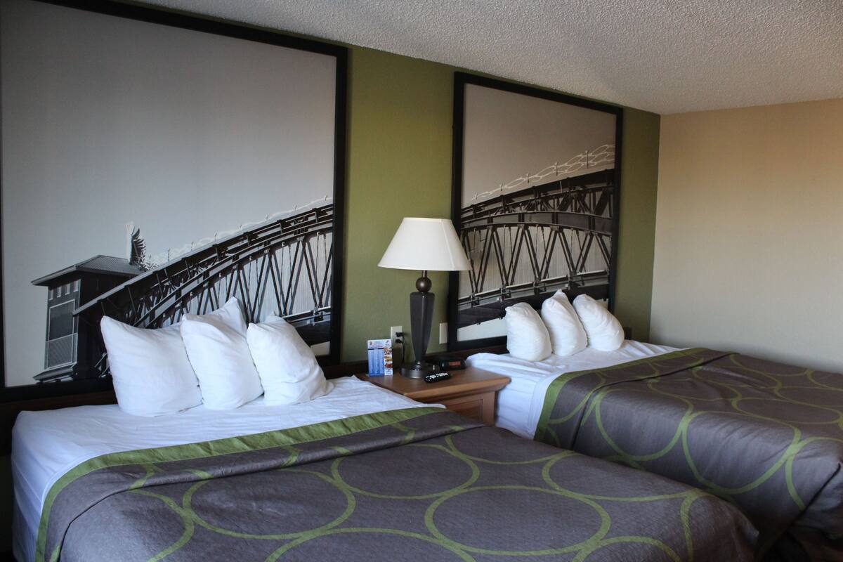 Quarto de hotel com duas camas, uma mesa de cabeceira com abajur e uma obra de arte na parede.