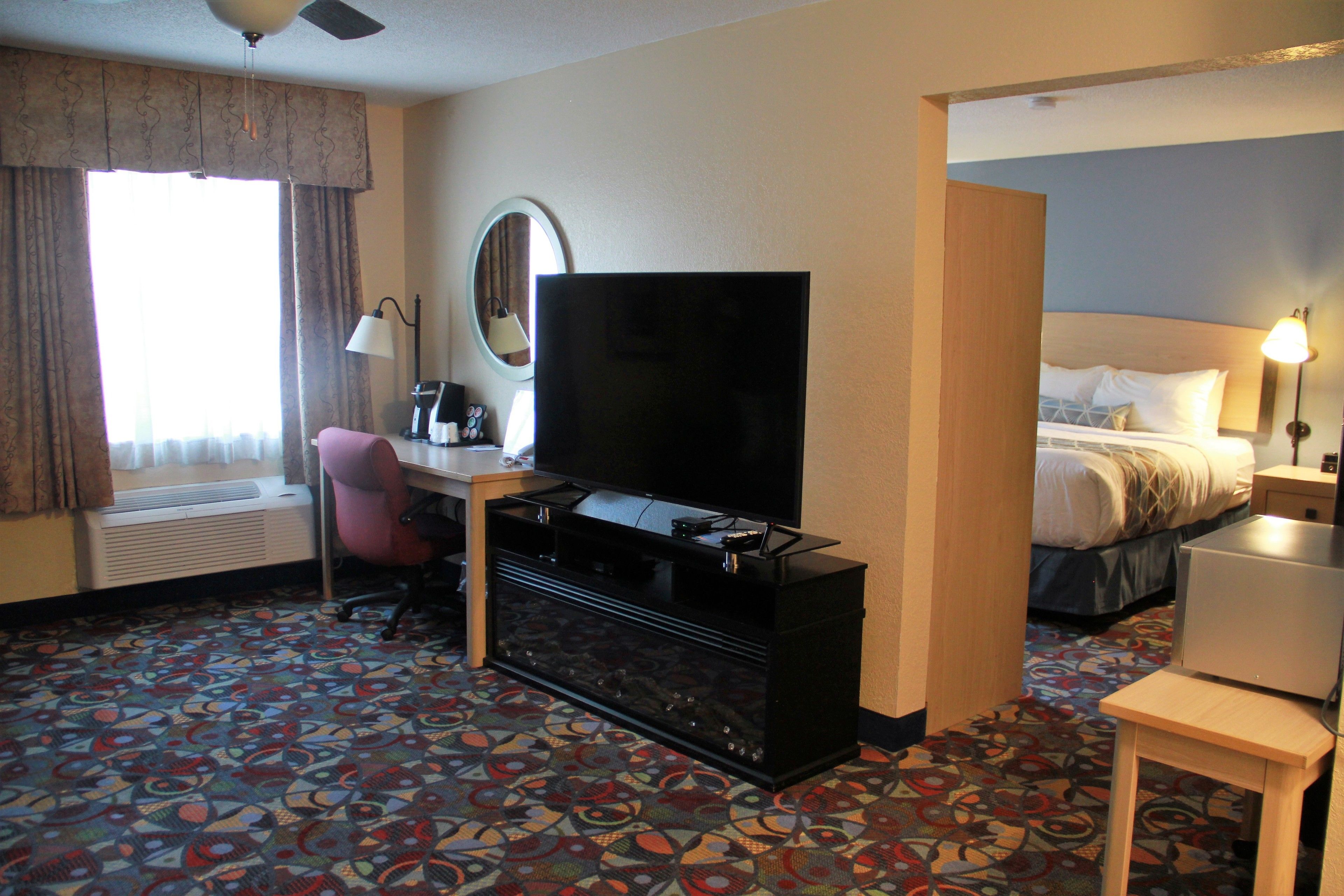 Quarto de hotel com uma televisão de tela grande, uma escrivaninha com abajur, uma cadeira, uma cama com cabeceira e um tapete estampado.