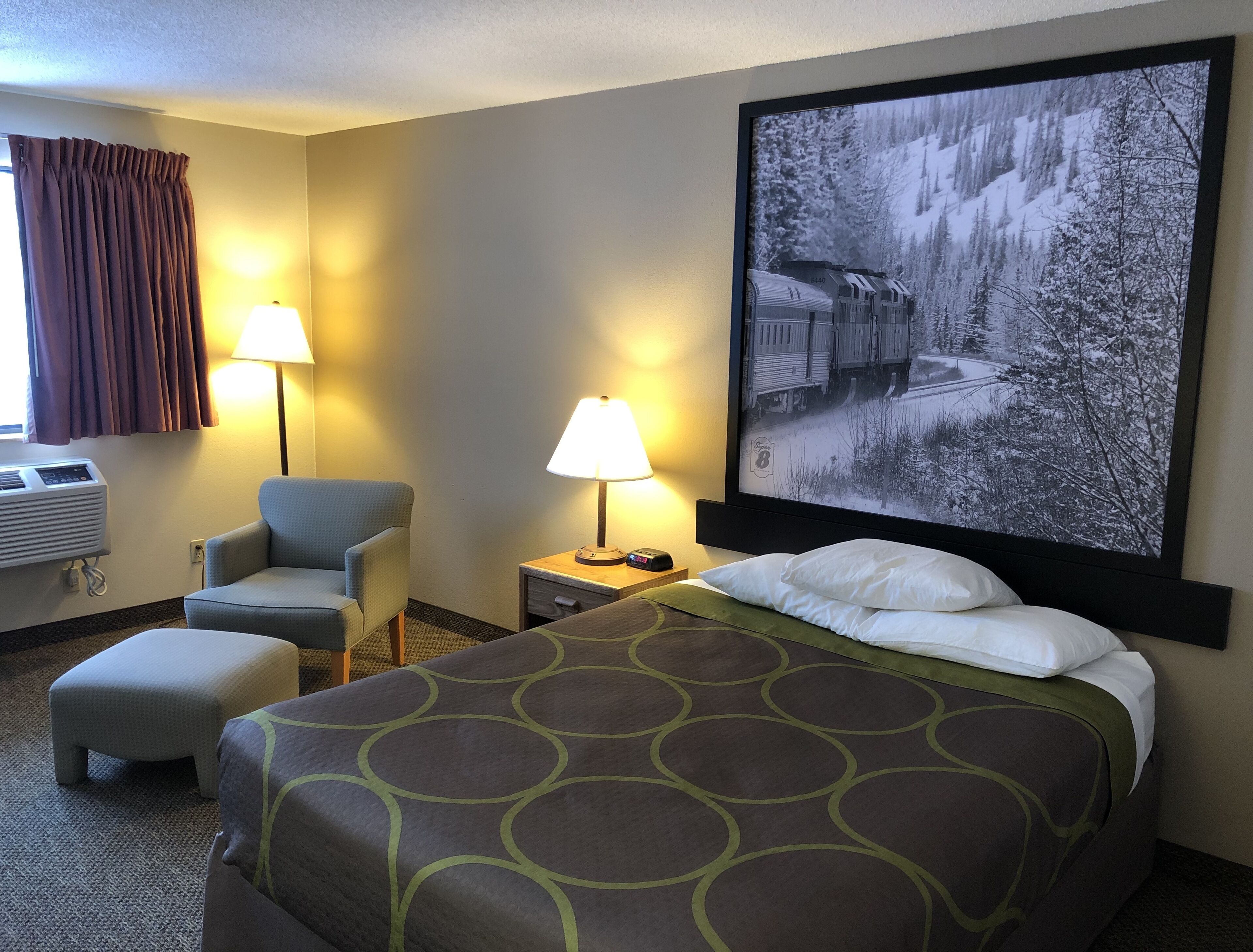 Quarto de hotel com uma cama, duas cadeiras, uma escrivaninha com uma luminária, ar condicionado e uma grande moldura na parede.