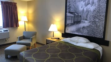 Quarto de hotel com uma cama, duas cadeiras, uma escrivaninha com uma luminária, ar condicionado e uma grande moldura na parede.