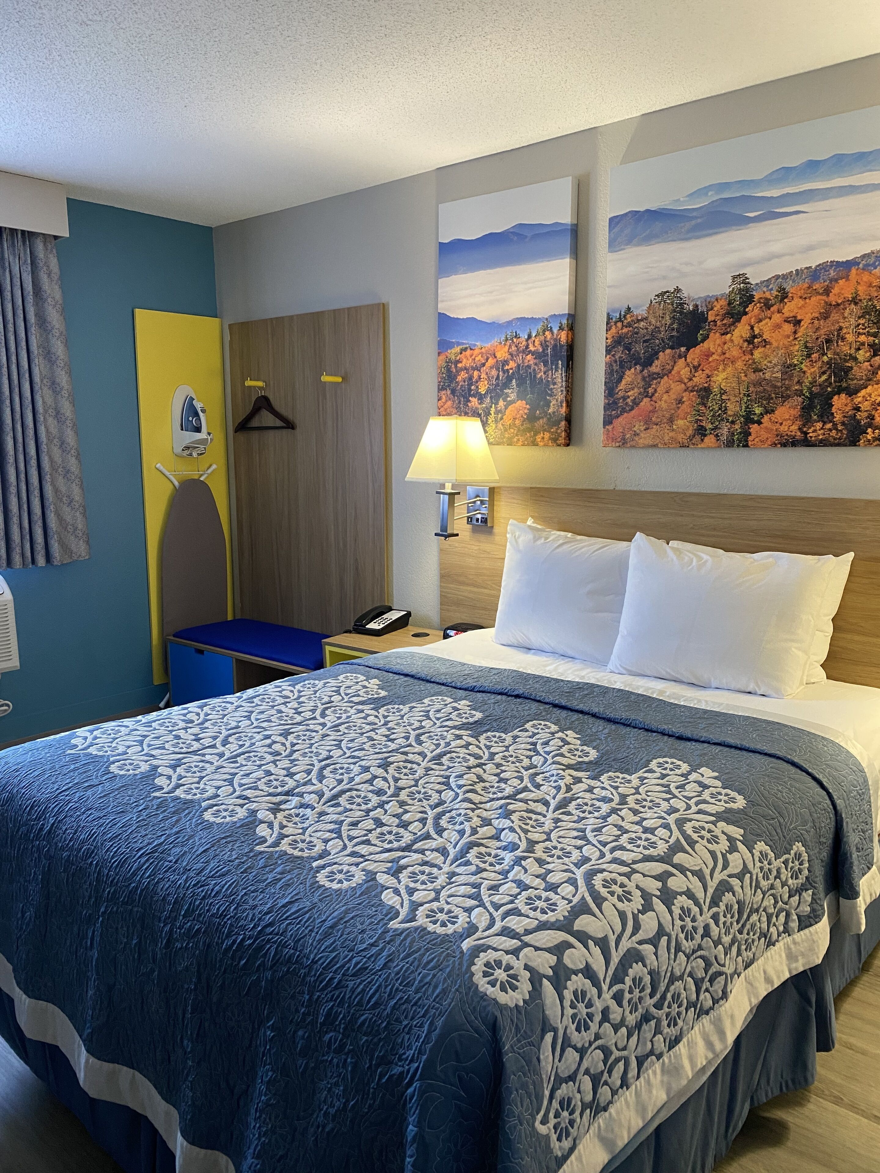 Quarto de hotel com uma cama grande, um quadro de folhagem de outono, uma escrivaninha com abajur e uma cadeira azul.