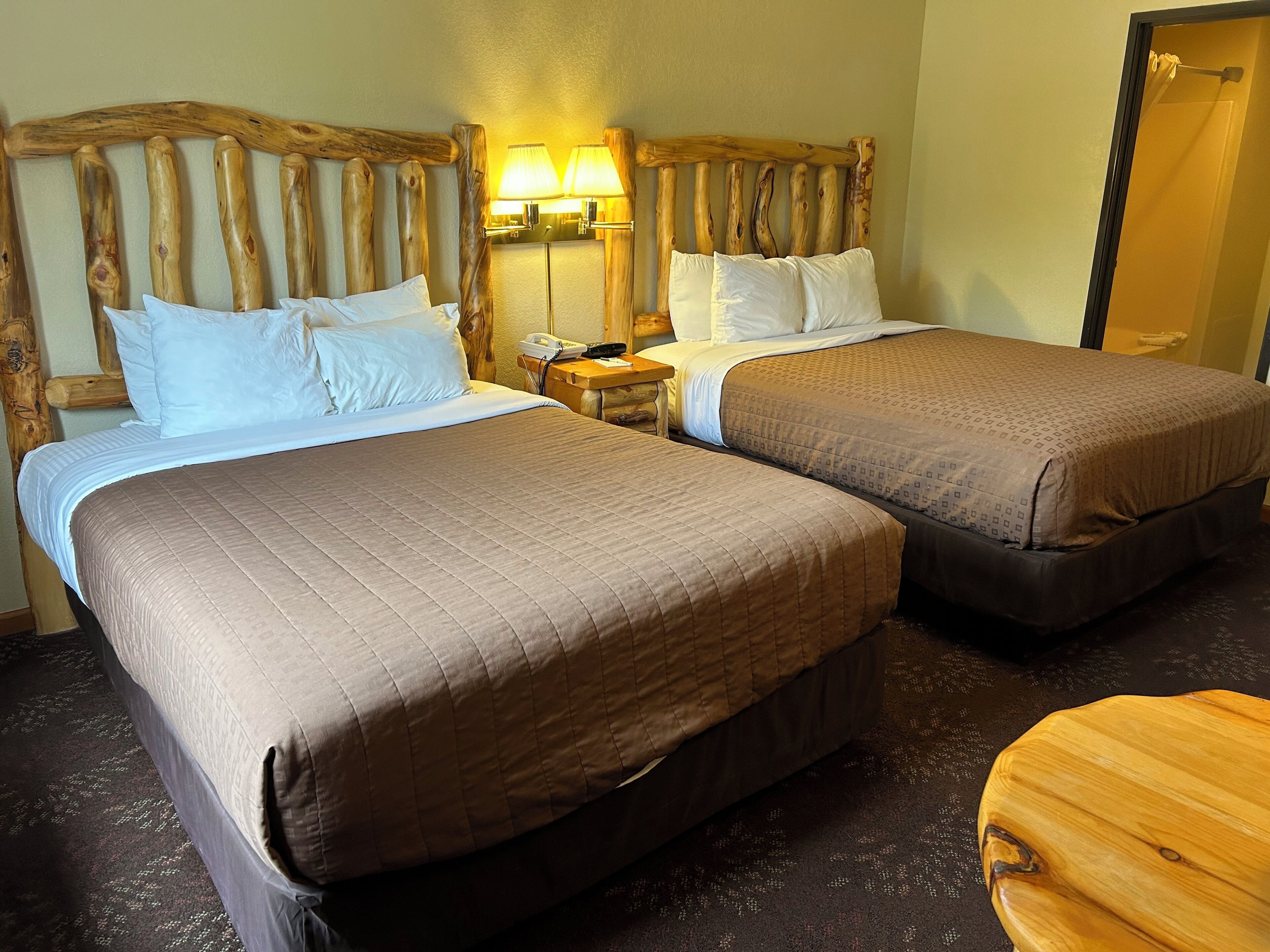 Quarto de hotel com duas camas, cabeceiras de madeira, mesa de cabeceira com abajur e um pequeno banco de madeira.