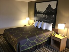 Quarto de hotel com uma cama grande, duas luminárias de cabeceira, uma mesinha de cabeceira e um quadro com paisagem de montanhas.