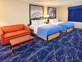 Quarto de hotel com duas camas, um sofá, uma mesa pequena e duas luminárias fixadas na parede.