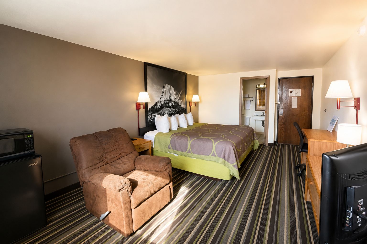 Quarto de hotel com cama, cadeira, escrivaninha, televisão e micro-ondas.