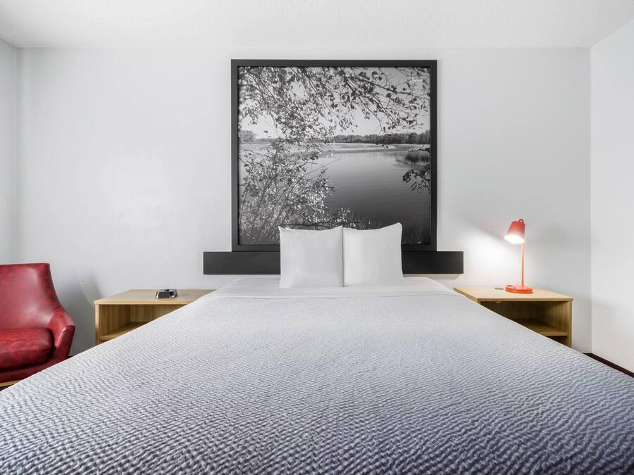 Quarto de hotel moderno com cama grande, uma cadeira vermelha, mesas de cabeceira em madeira e uma luminária fixada na parede.