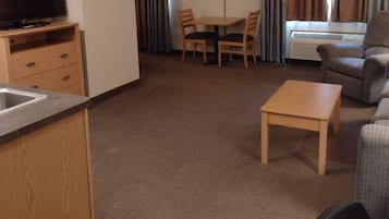 Quarto de hotel compacto com cozinha compacta, televisão de tela plana, área de jantar com mesa e cadeiras, sofá e janela com cortinas.