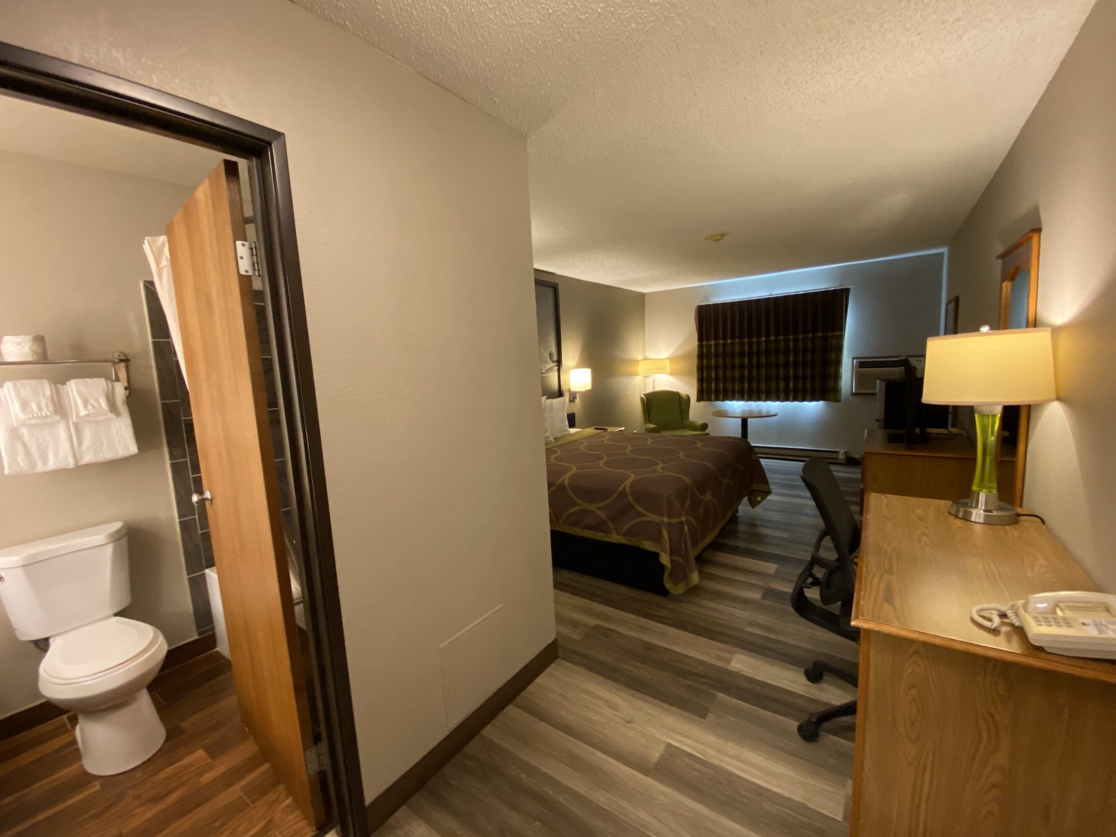 Quarto de hotel com piso de madeira, uma cama com colcha marrom, uma escrivaninha com luminária, telefone e uma cadeira.