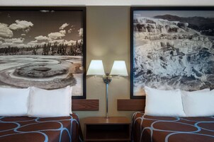 Quarto de hotel com duas camas, uma mesinha de cabeceira e uma luminária. Há dois grandes murais na parede retratando paisagens naturais.