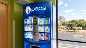 Máquina automática azul da Pepsi com diversas opções de bebidas, em frente a uma parede verde.