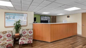 Uma área de recepção com um balcão de madeira, duas cadeiras estampadas, uma pequena mesa com uma planta e um mural com diversos avisos.