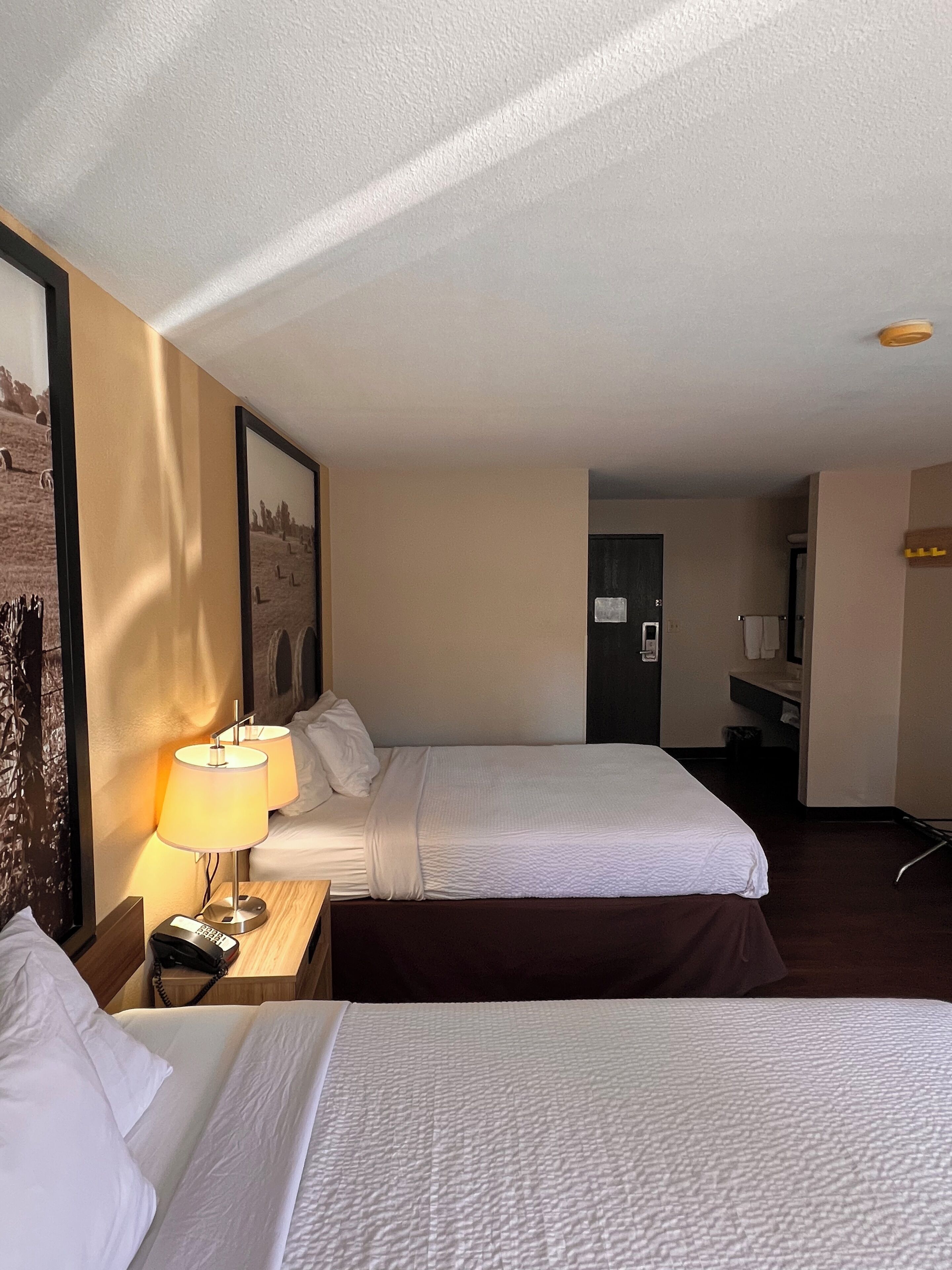 Quarto de hotel com duas camas, uma mesa de cabeceira com luminária, um quadro na parede e um banheiro visível por uma porta aberta.