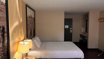 Quarto de hotel com duas camas, uma mesa de cabeceira com luminária, um quadro na parede e um banheiro visível por uma porta aberta.