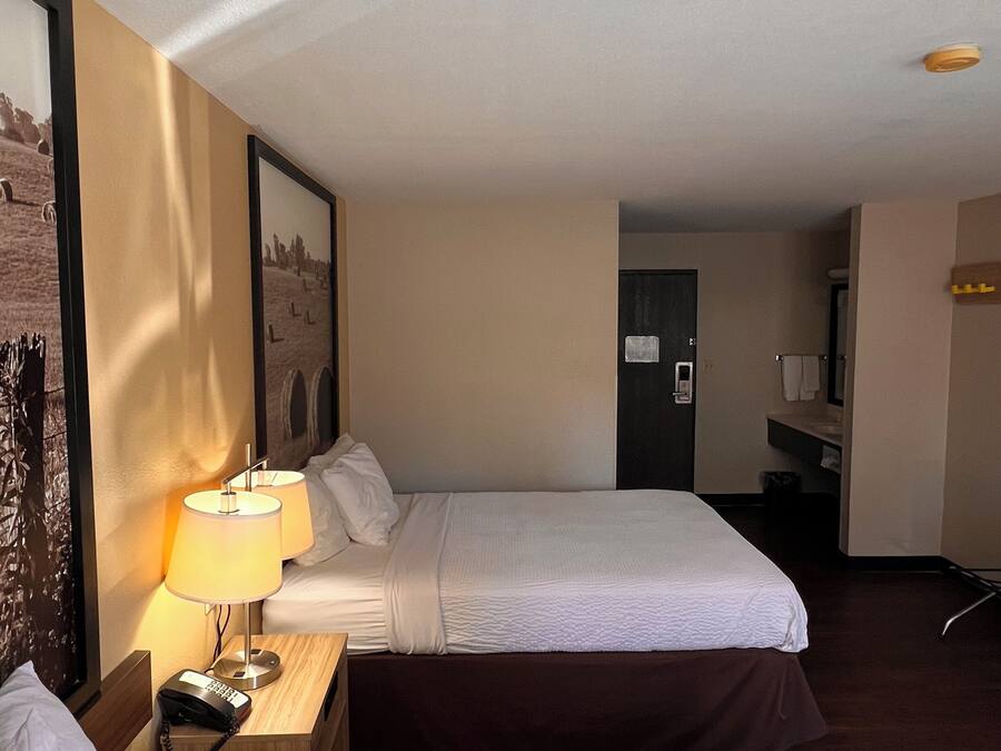 Quarto de hotel com duas camas, uma mesa de cabeceira com luminária, um quadro na parede e um banheiro visível por uma porta aberta.