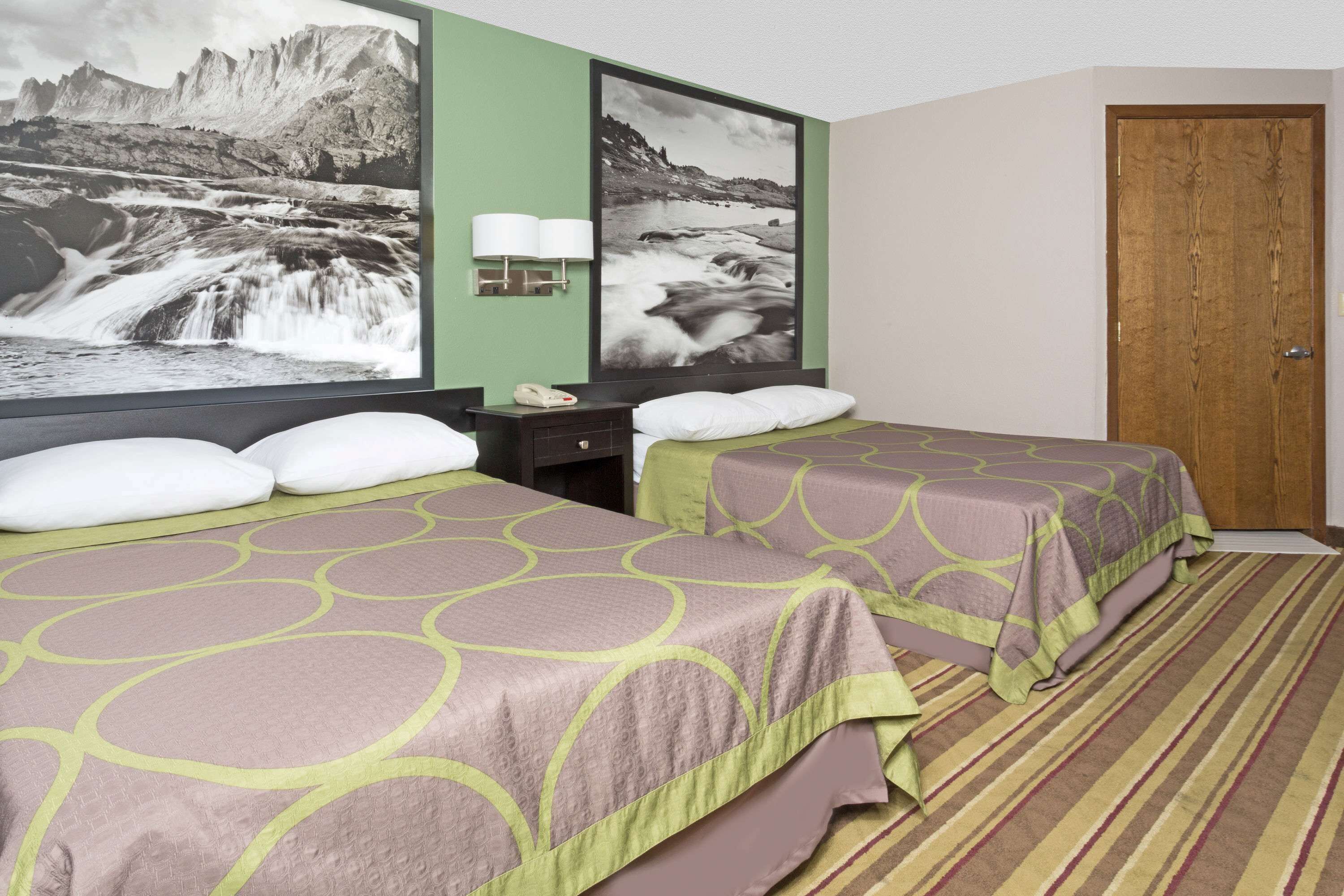 Quarto de hotel com duas camas, uma fotografia grande emoldurada na parede, uma porta de madeira e um tapete listrado.