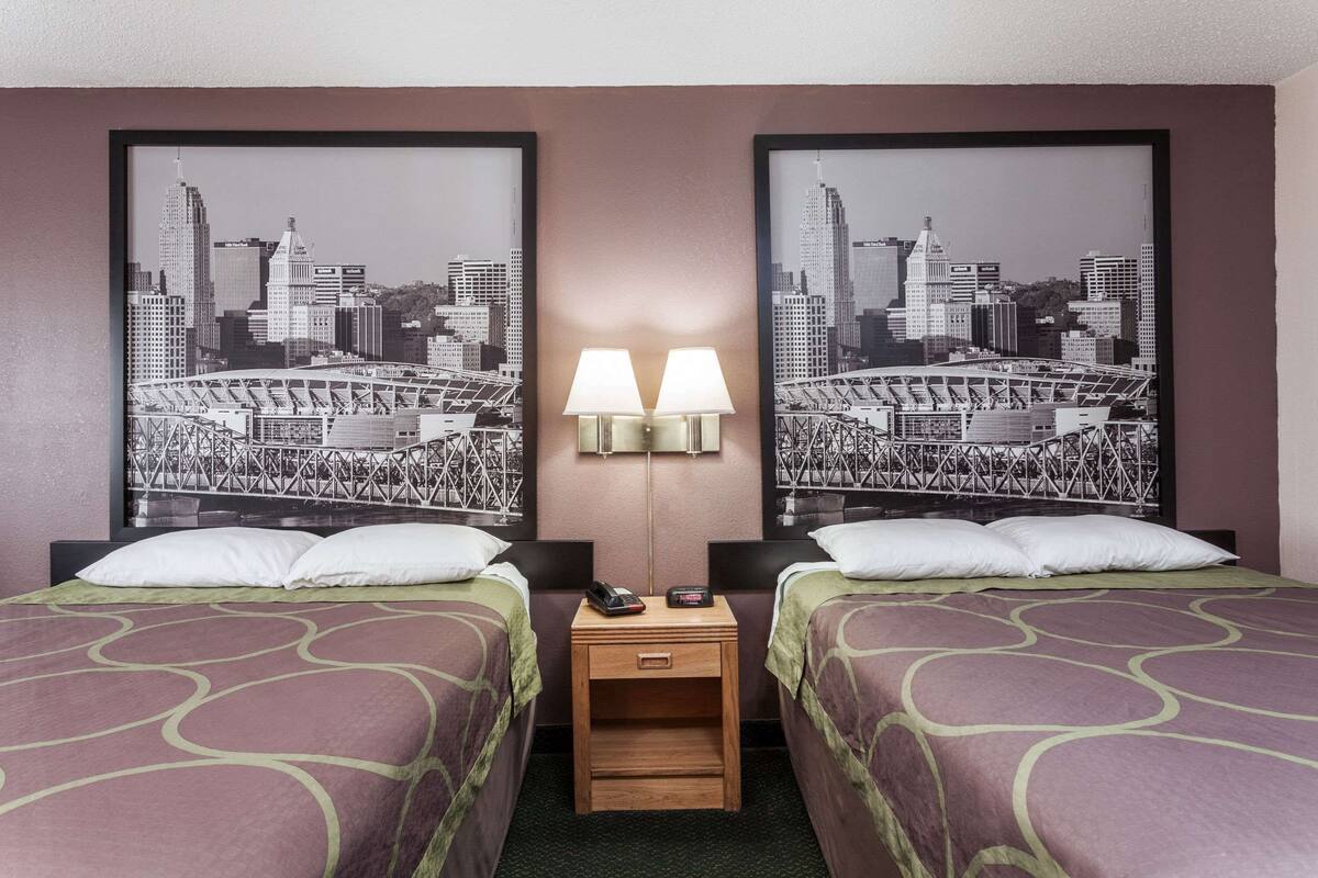 Quarto de hotel com duas camas, uma mesa de cabeceira com abajur e duas imagens emolduradas com vista urbana na parede.
