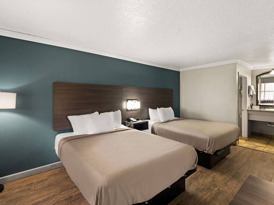 Quarto de hotel com duas camas, uma mesa de cabeceira, uma luminária, um espelho e um banheiro com pia e espelho.