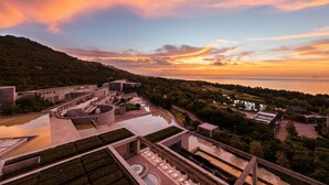 Um complexo hoteleiro moderno com uma piscina grande, áreas de estar ao ar livre e uma vista panorâmica para a água e colinas ao pôr do sol.