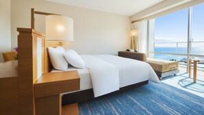 Quarto de hotel com uma cama grande, uma mesa de cabeceira, uma luminária, uma mesinha de madeira e vista para o oceano através de uma janela ampla.