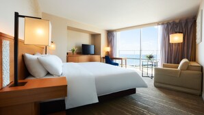 Quarto de hotel com cama grande, escrivaninha com televisão, uma cadeira e vista para o oceano.