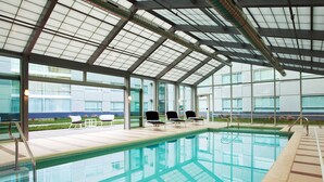 Piscina interior com paredes de vidro transparente, cadeiras de descanso e vista para o exterior do edifício.
