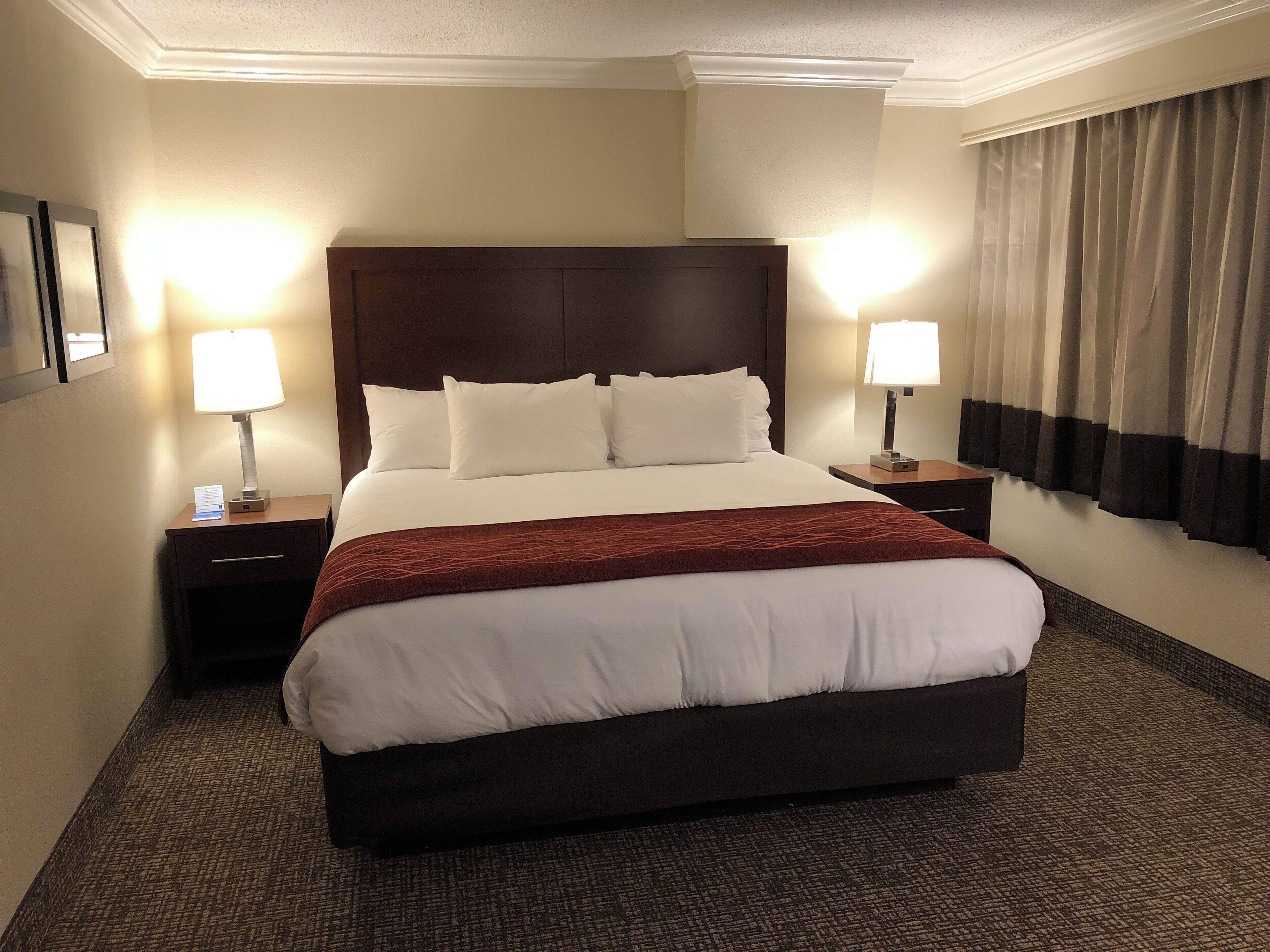 Quarto de hotel com uma cama grande, duas mesas de cabeceira com abajures, um espaldar escuro e uma janela com cortinas.