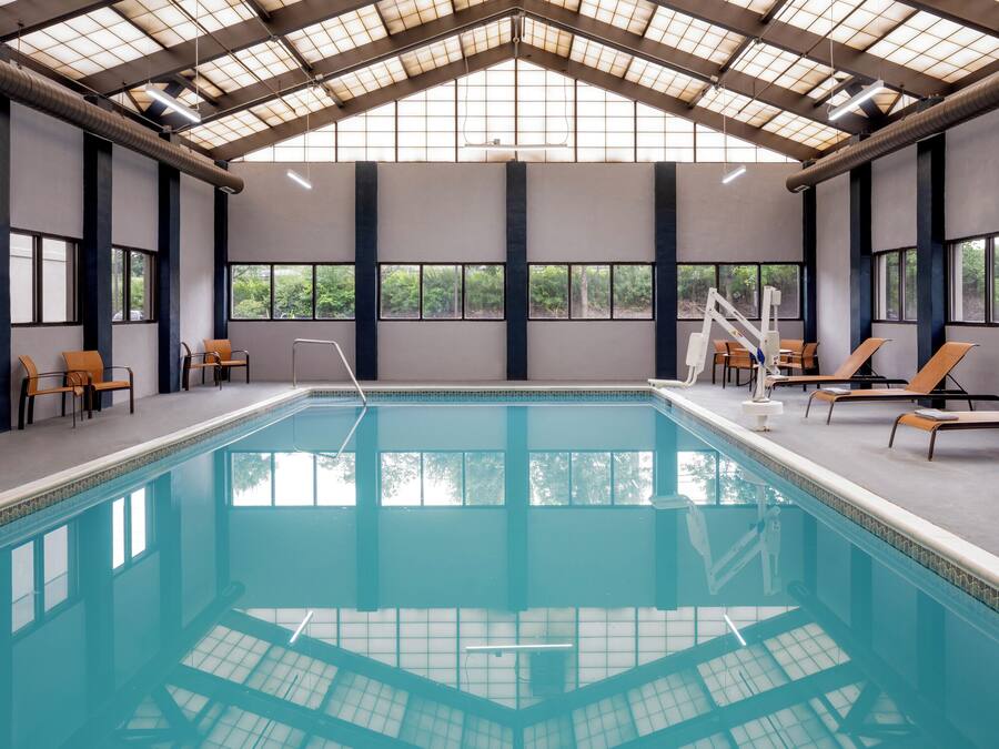 Piscina interna com água azul cristalina, rodeada por espreguiçadeiras e um teto alto com claraboias.
