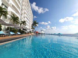 Uma piscina azul cristalina com espreguiçadeiras e guarda-sóis, um edifício branco com varandas, e vista para o mar.