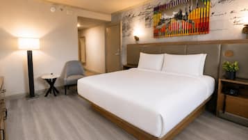 Quarto de hotel com uma cama grande, mesa de cabeceira com abajur, uma cadeira e parede com pintura abstrata colorida.