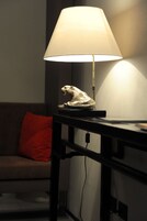 Um abajur com cúpula branca sobre uma mesa preta, um travesseiro vermelho em um sofá e uma tomada na parede.