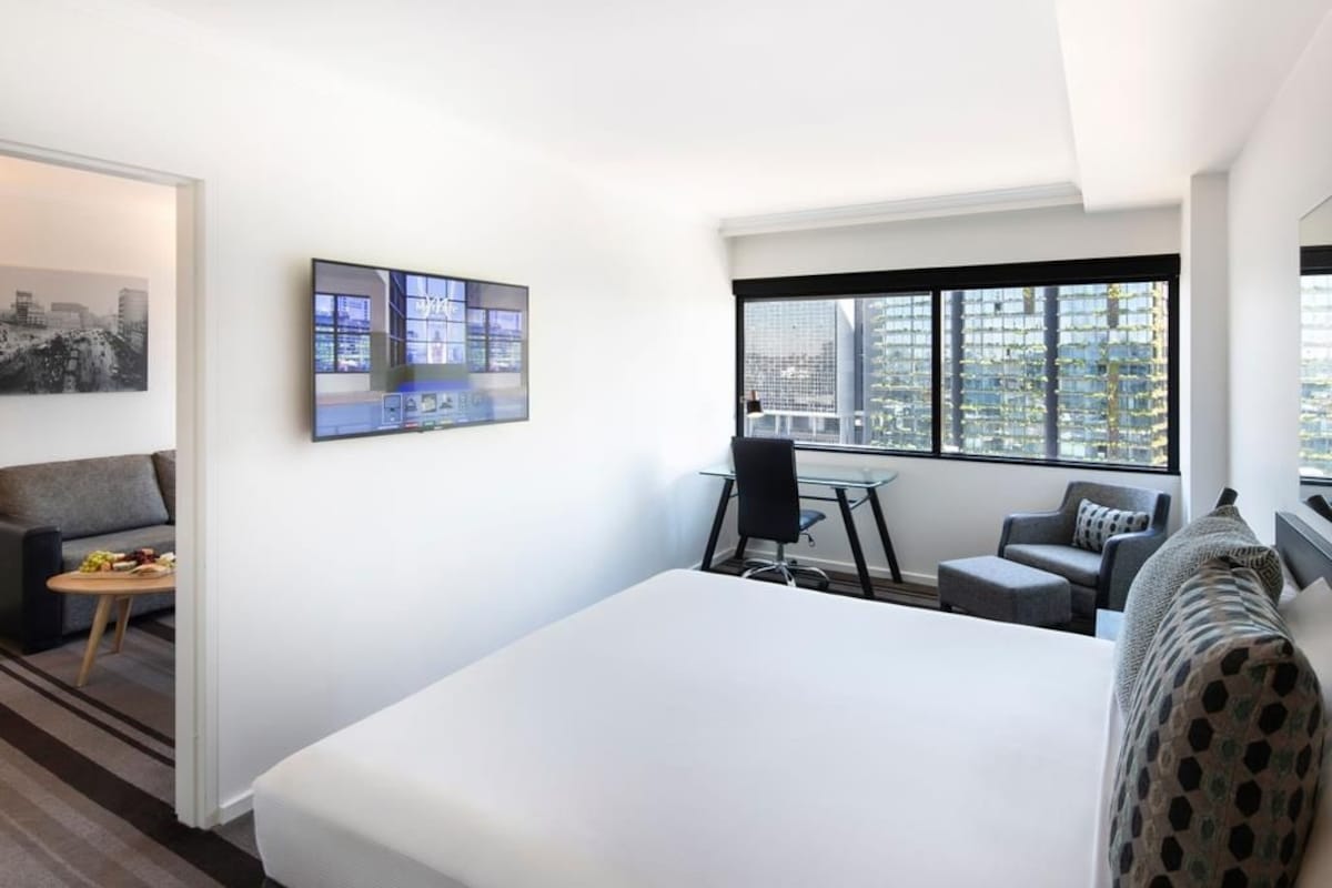 Quarto de hotel moderno com uma cama grande, uma escrivaninha, uma cadeira, um sofá e uma janela ampla com vista para a cidade.