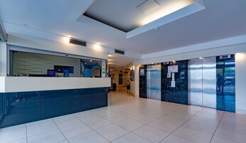 Uma área de recepção de hotel, com balcão de atendimento, bebedouro e elevador.