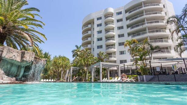 Um hotel com piscina, palmeiras e uma cascata.