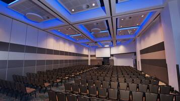 Sala de conferências com fileiras de cadeiras, uma tela grande e iluminação azul no teto.