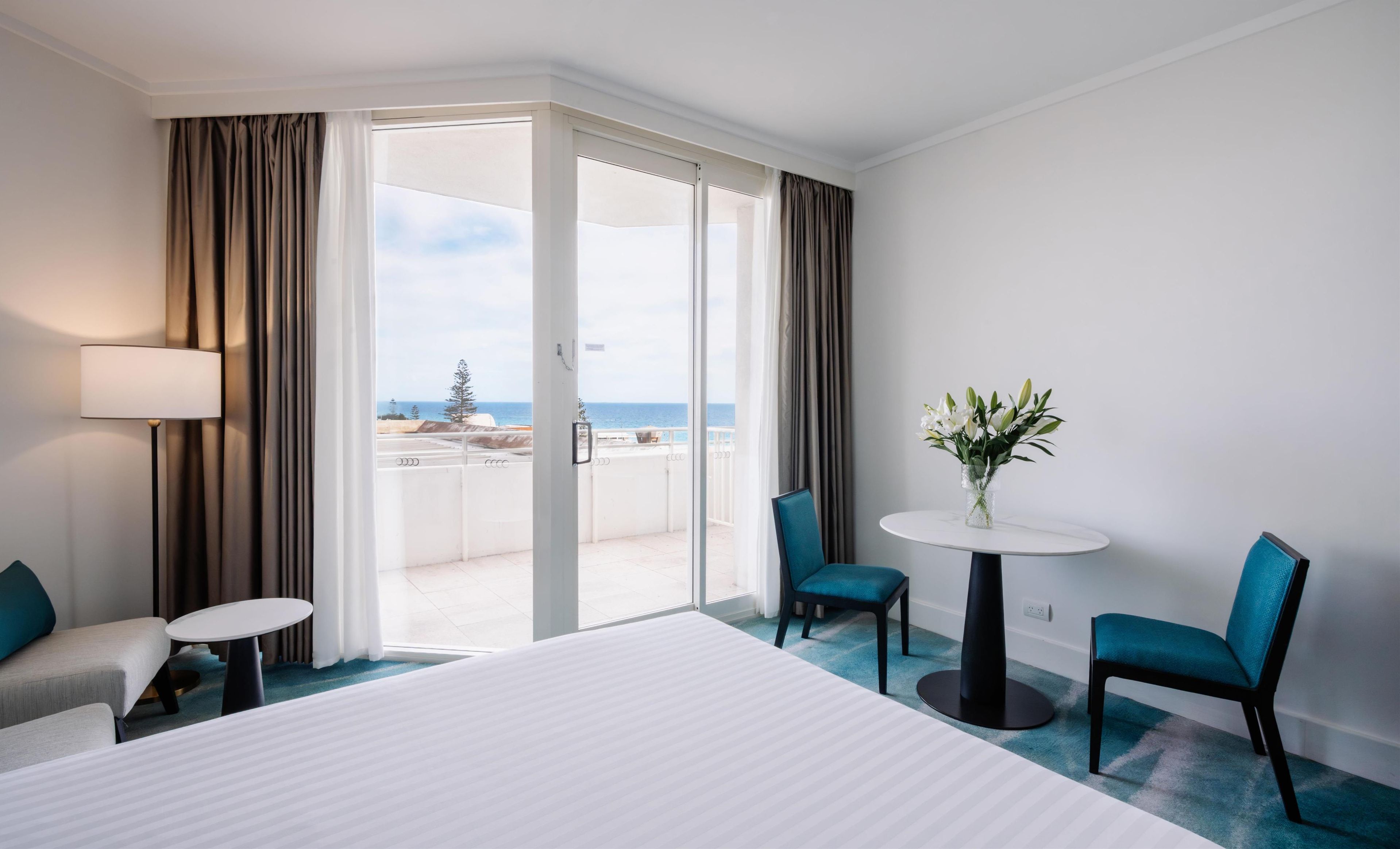 Quarto de hotel com uma cama grande, duas cadeiras, uma mesinha e varanda com vista para o mar.