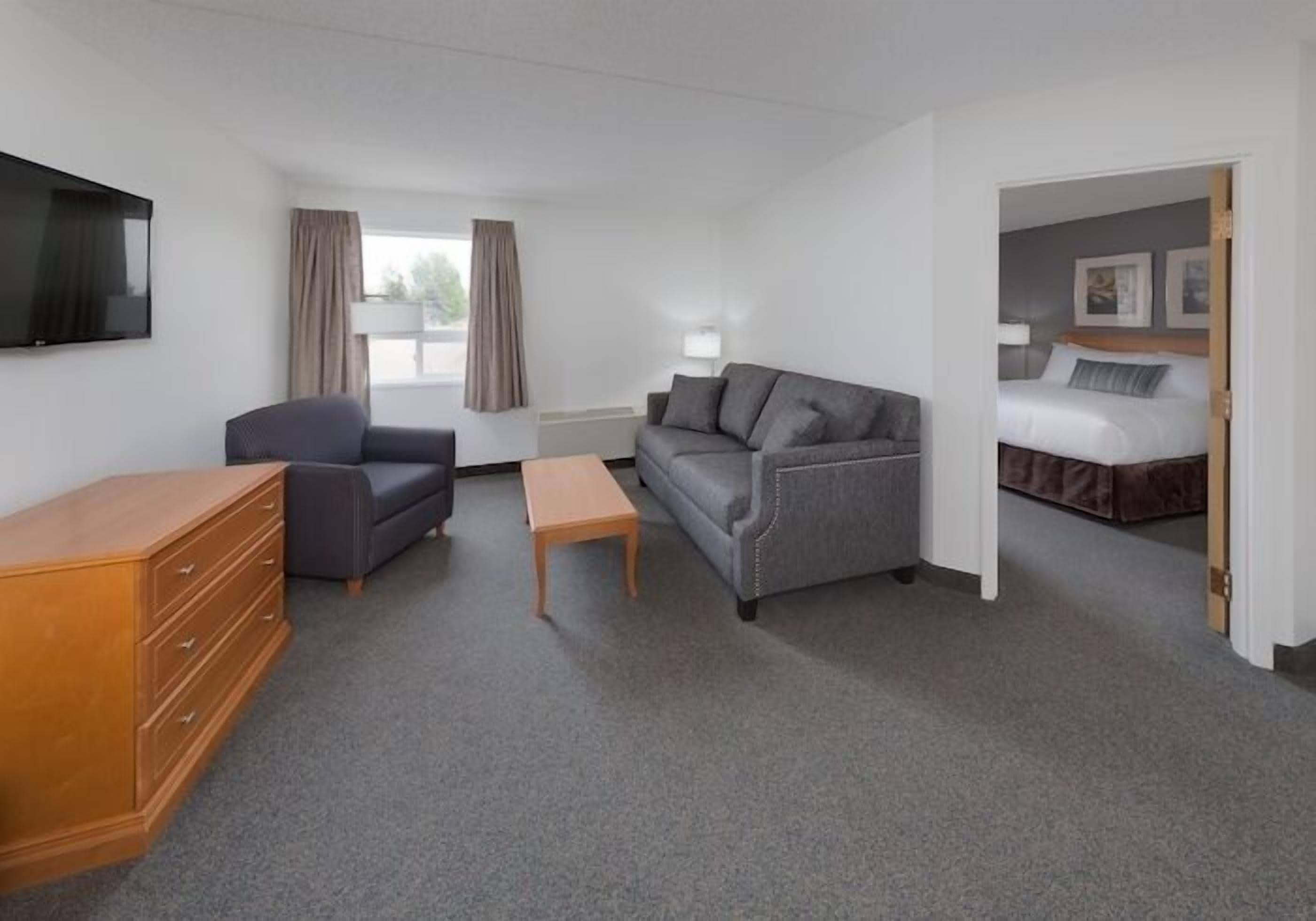 Quarto de hotel com sofá cinzento, cómoda de madeira, mesa pequena, televisão na parede e cama com cabeceira.