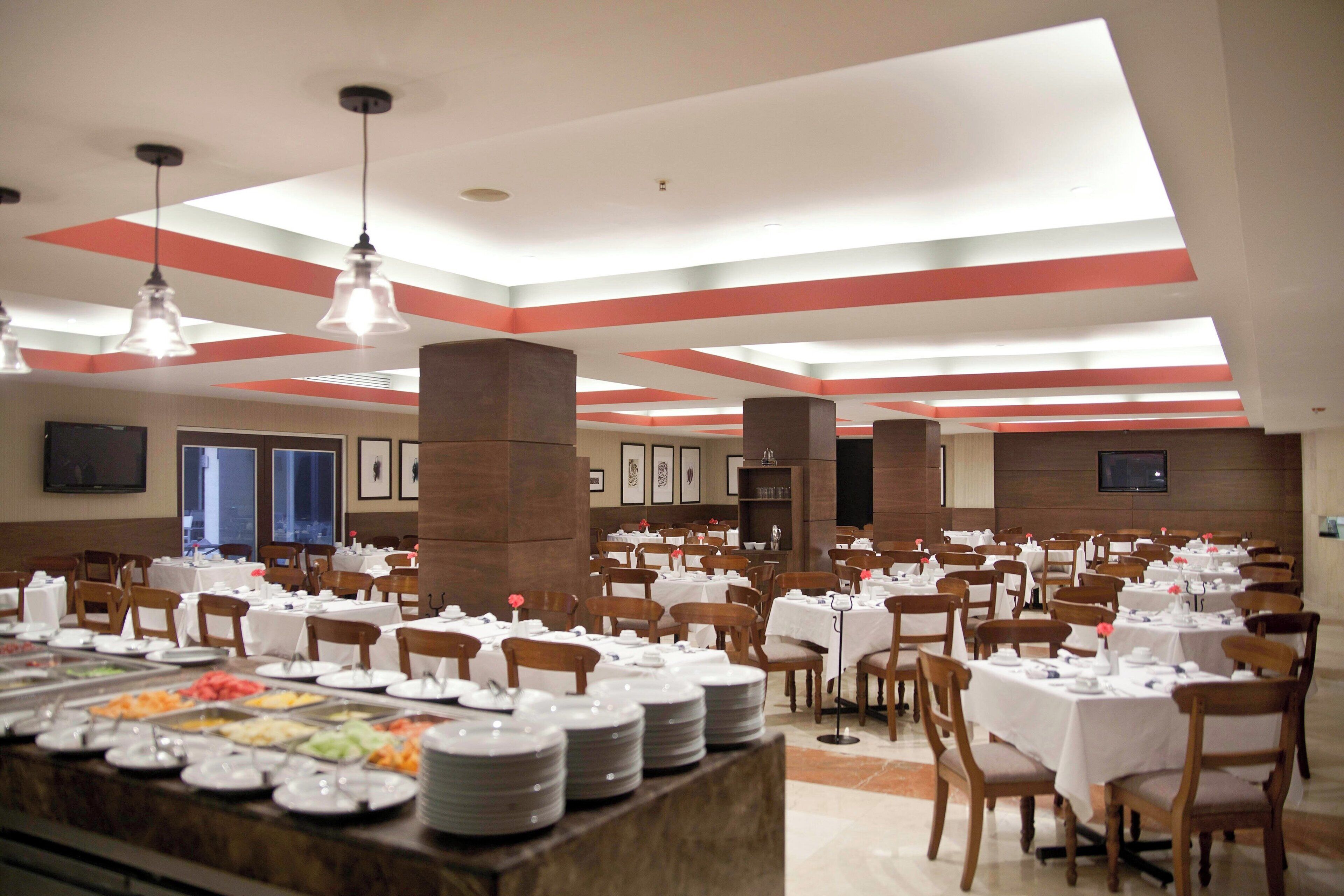 Um amplo salão de refeições com diversas mesas e cadeiras, um espaço de buffet com várias opções de comida e um teto alto com detalhes em vermelho e branco.
