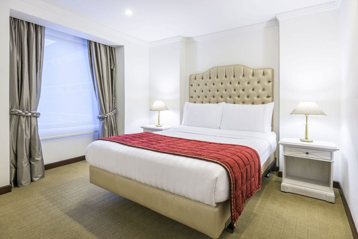 Quarto de hotel com uma cama grande, duas mesas de cabeceira, uma janela com cortinas e um chão claro.