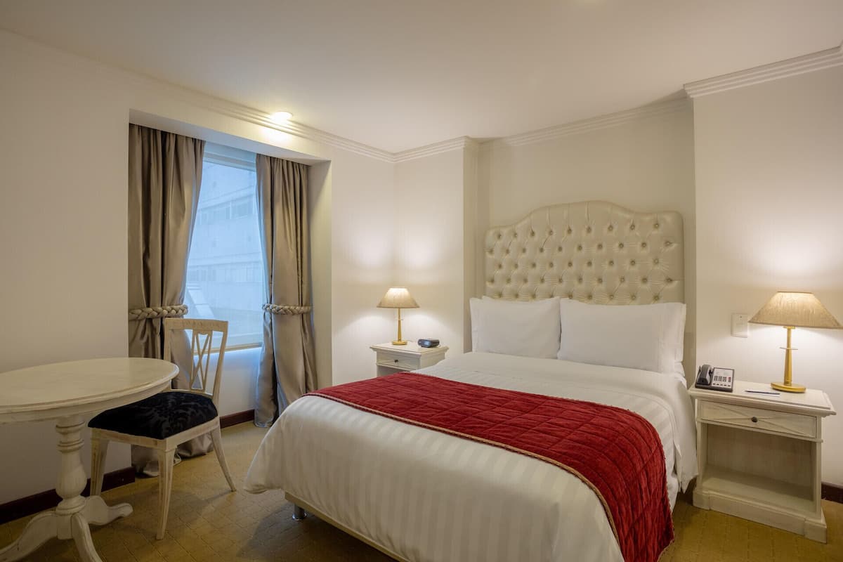 Quarto de hotel com uma cama, duas mesas de cabeceira, uma cadeira, uma mesinha e uma janela com cortinas.