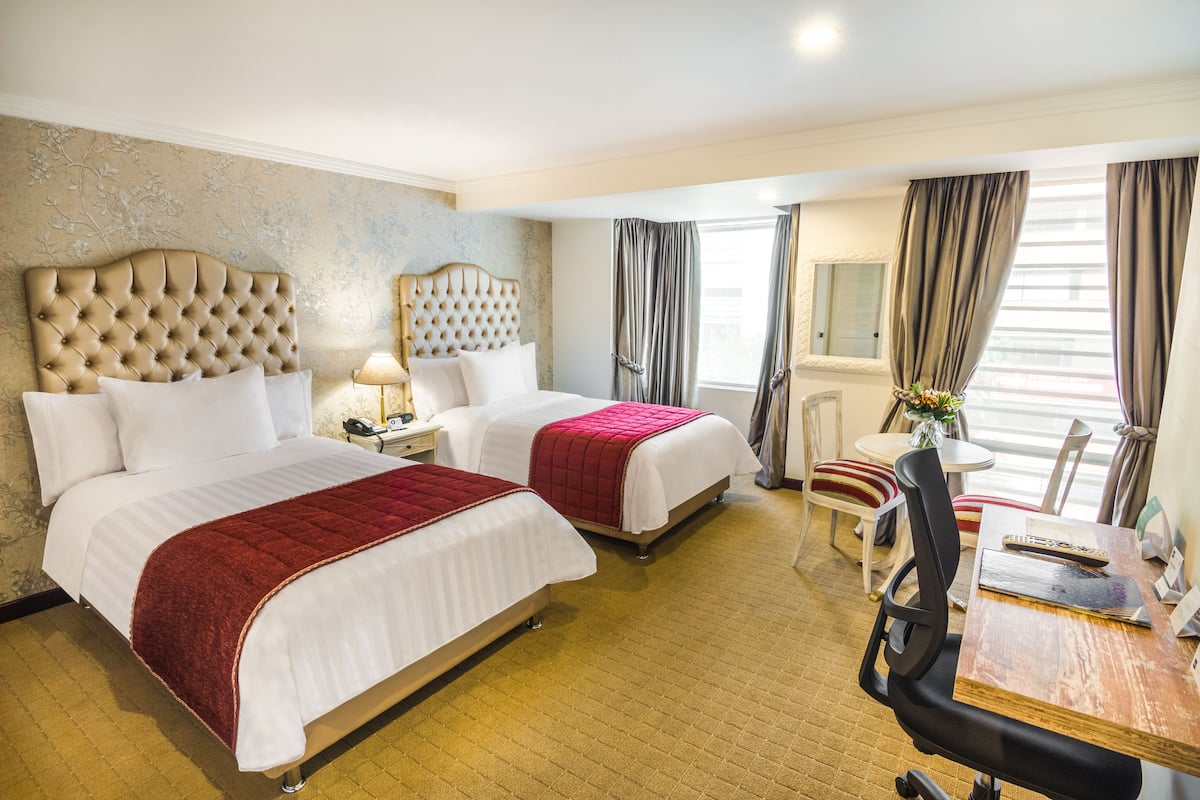 Quarto de hotel com duas camas, uma escrivaninha, uma cadeira, um abajur e um espelho.
