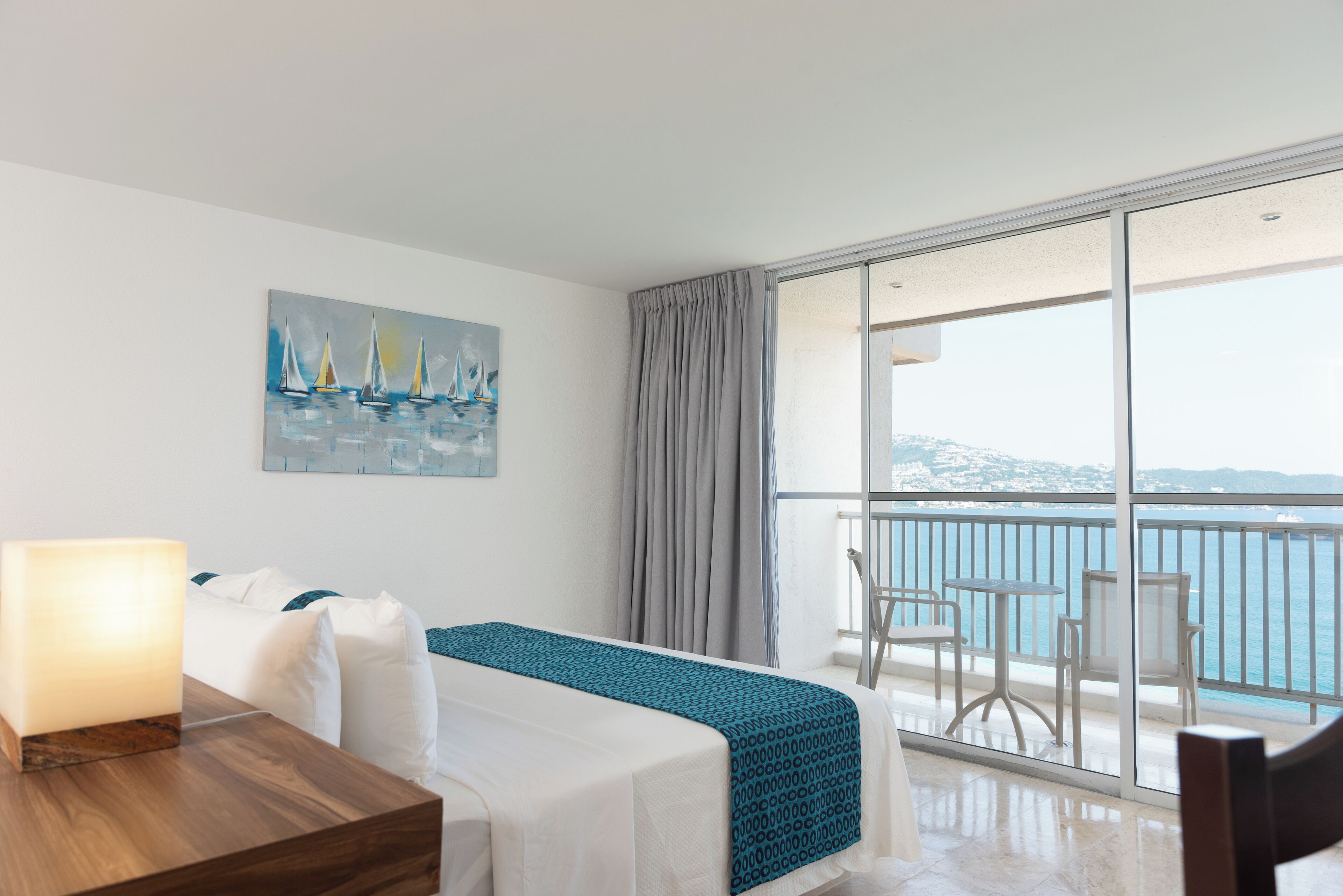 Quarto de hotel com uma cama, mesa de cabeceira com abajur, varanda com vista para o mar e um quadro na parede.