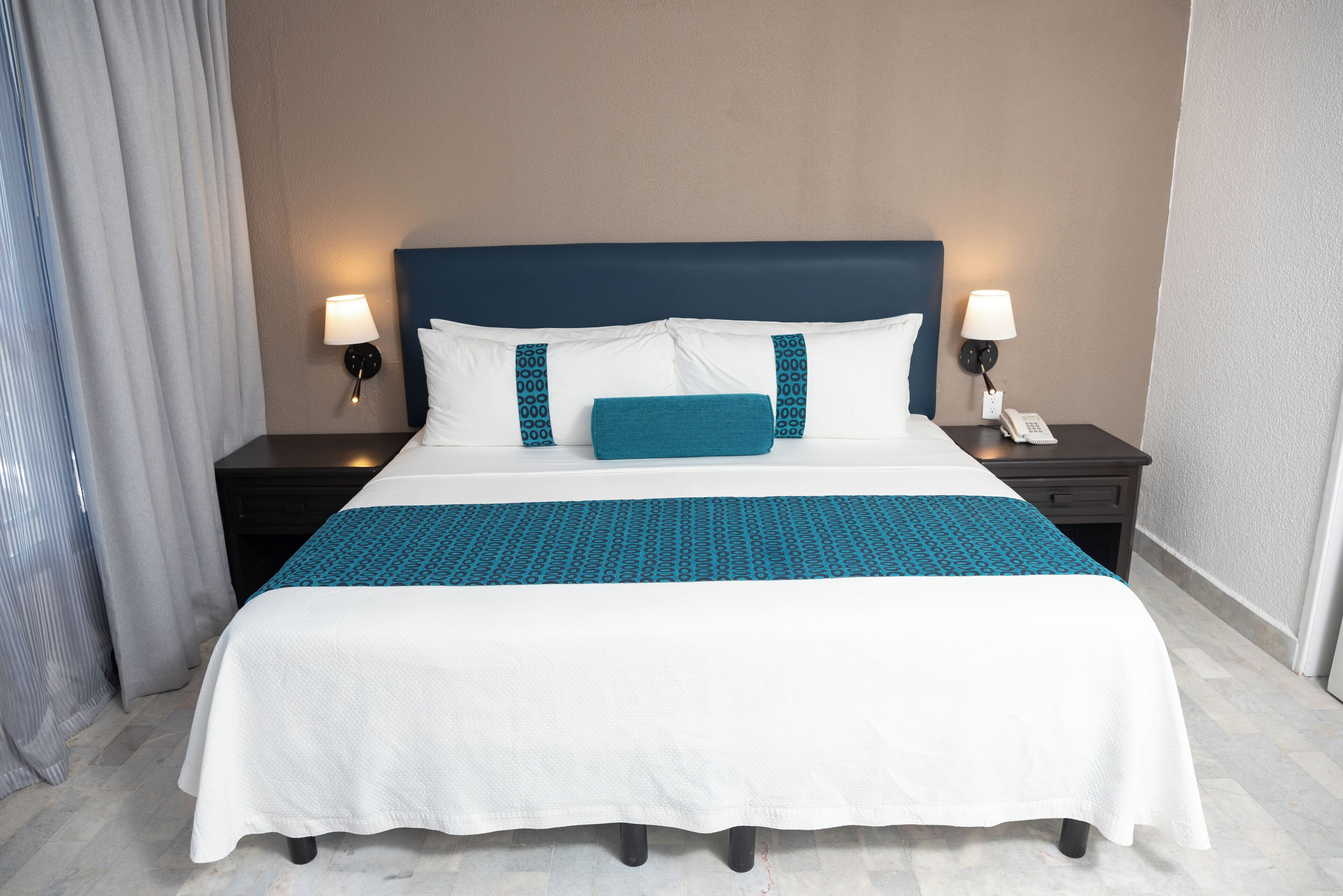 Uma cama bem arrumada com colcha branca e azul, duas almofadas combinando e uma almofada de destaque azul. Há duas mesas de cabeceira, uma com abajures e um telefone.