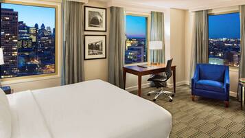 Quarto de hotel com uma cama grande, uma escrivaninha com cadeira, uma poltrona azul e vista para a cidade ao entardecer.