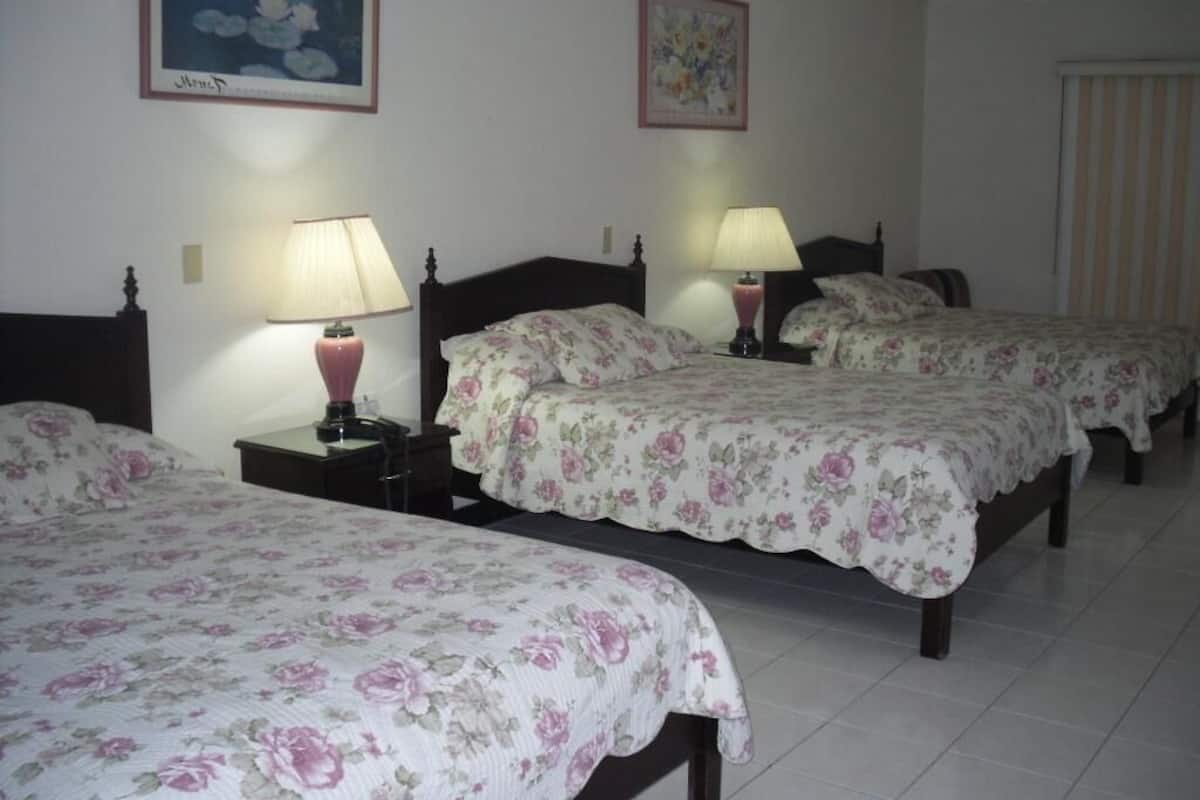 Quarto de hotel com três camas, cada uma com colcha florida, luminárias laterais e quadros nas paredes.