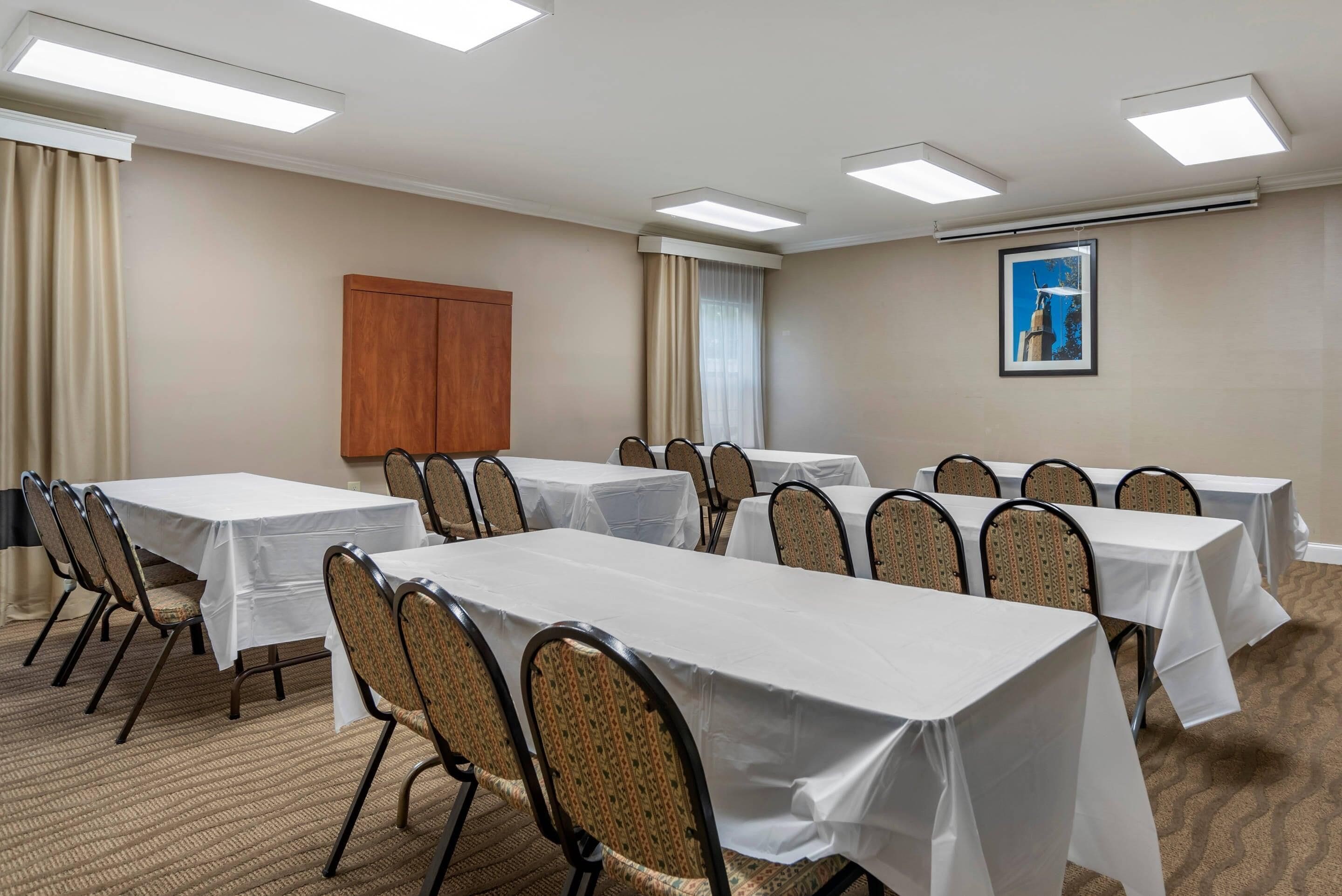 Uma sala de conferências com várias mesas e cadeiras, um armário de madeira e um quadro na parede.