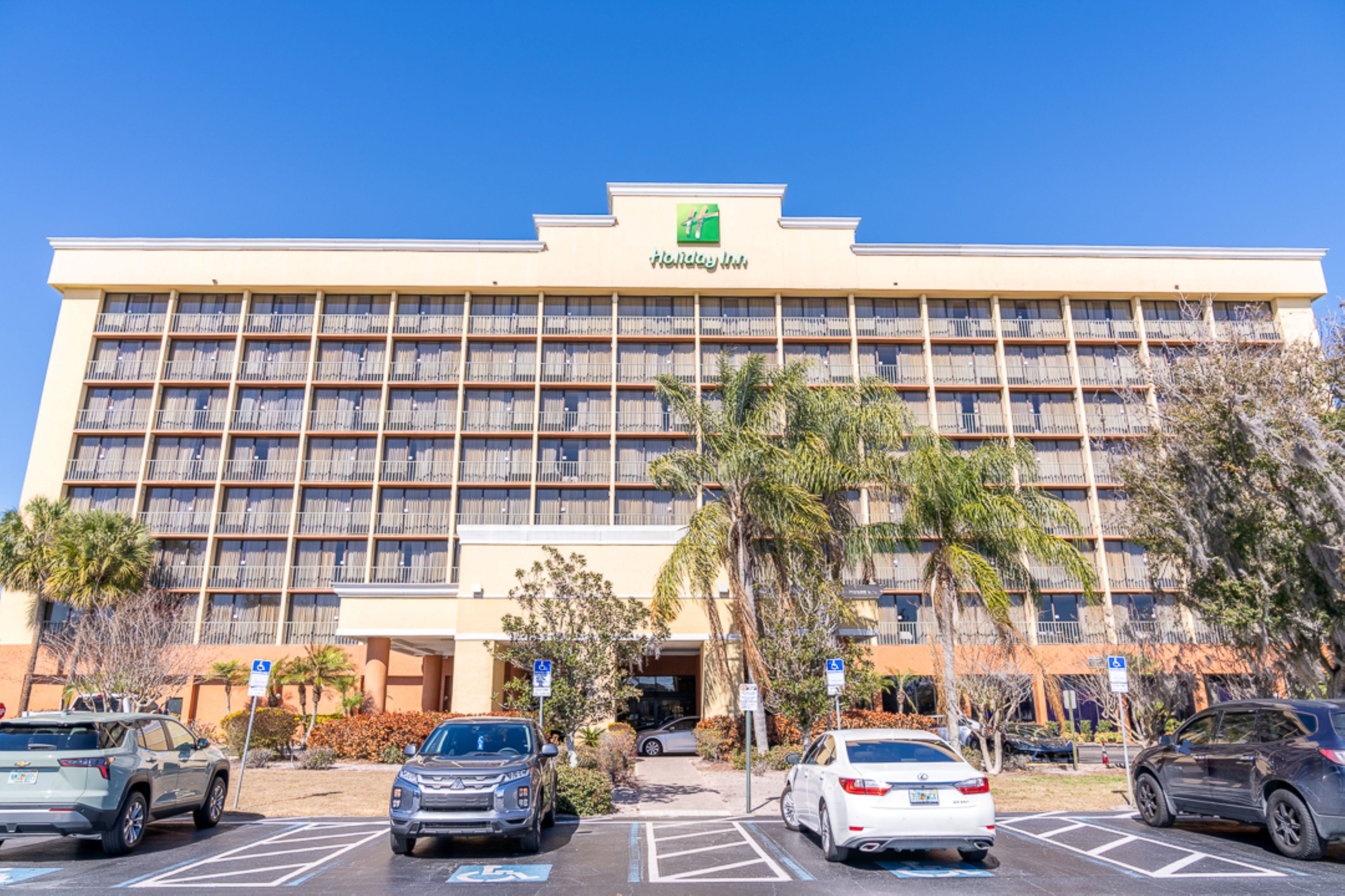 Um hotel Holiday Inn com um edifício de vários andares, estacionamento e vários carros estacionados em frente.